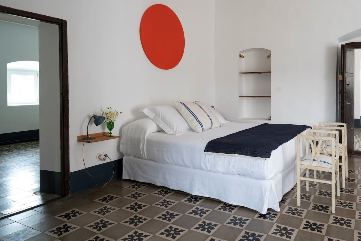 Quarto com cama, um ornamento circular vermelho na parede, mesa de cabeceira com abajur, cadeira e estante.