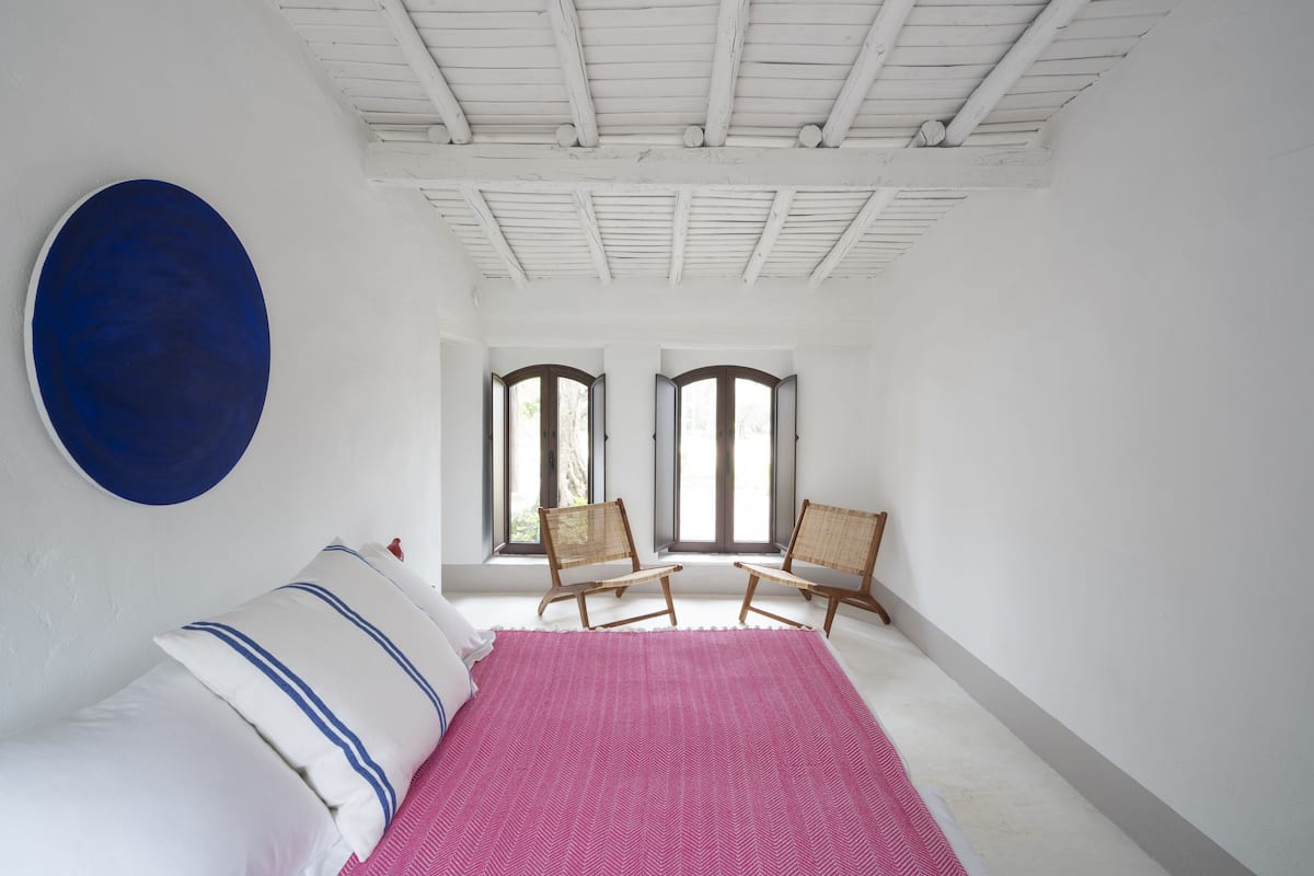 Quarto com cama, duas cadeiras de vime, uma grande obra de arte azul e duas janelas com molduras de madeira.