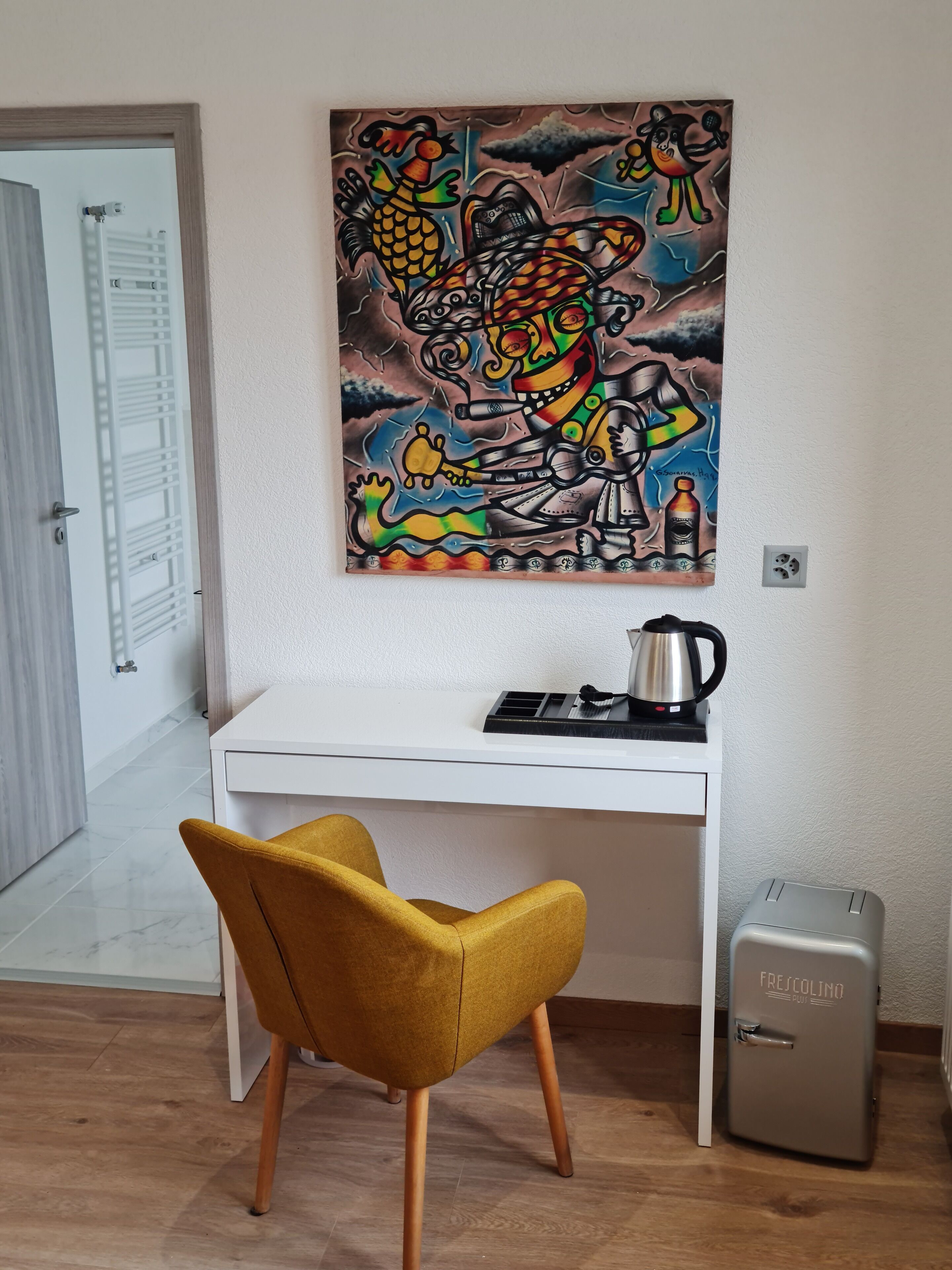 Quarto com escrivaninha branca, cadeira cor mostarda, pintura abstrata colorida, chaleira e pequeno frigorífico.