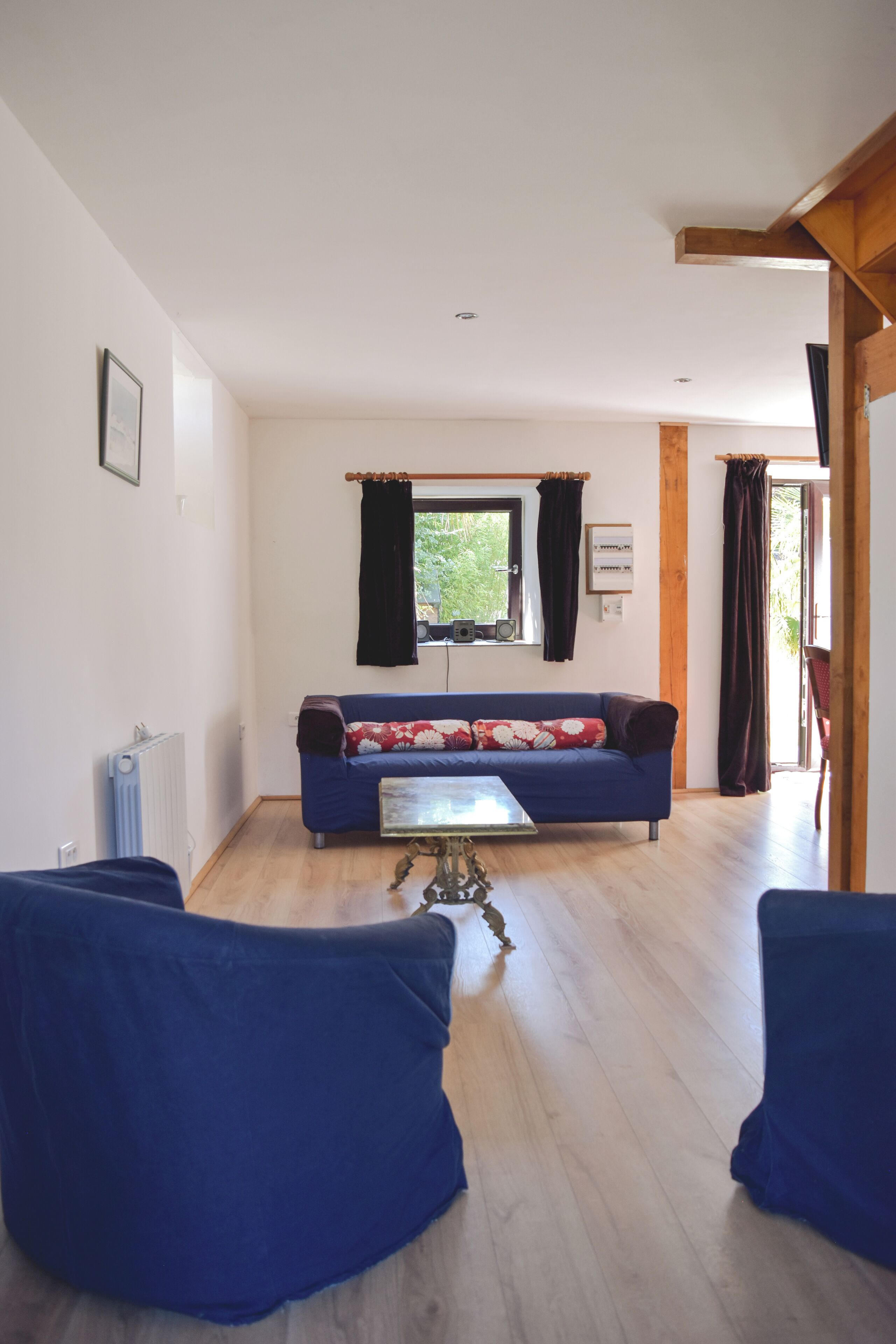 Quarto com um sofá azul, uma pequena mesa e duas cadeiras azuis. Há uma janela com cortinas, um radiador e uma viga de madeira no teto.