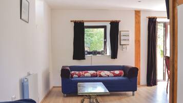 Quarto com um sofá azul, uma pequena mesa e duas cadeiras azuis. Há uma janela com cortinas, um radiador e uma viga de madeira no teto.