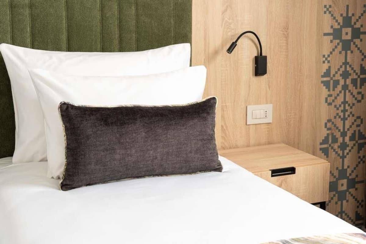 Cama com travesseiros brancos e cinza escuro, cabeceira verde, mesa de cabeceira de madeira com luminária preta fixada na parede e parede com padrão.