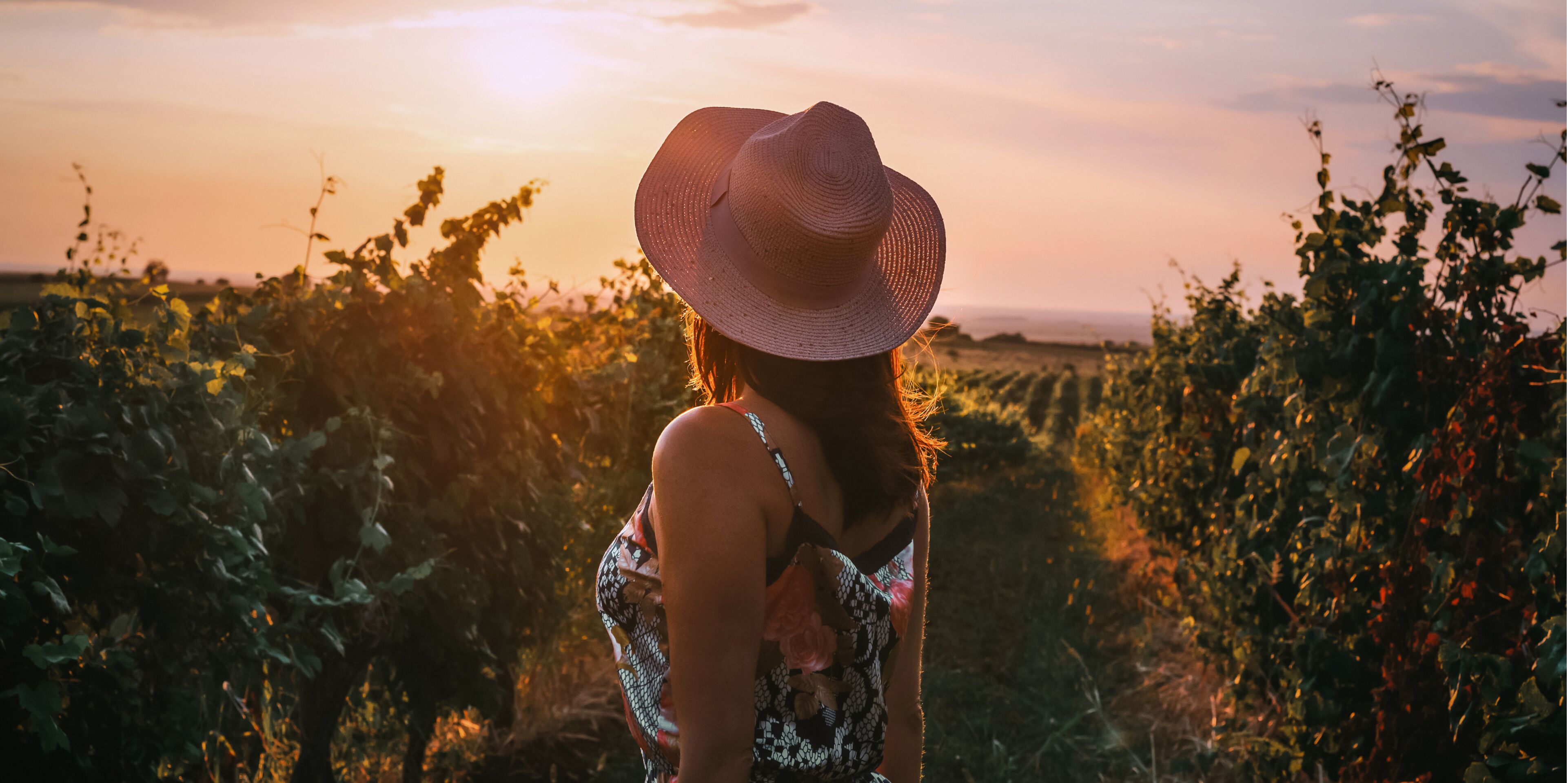 Uma pessoa de chapéu e vestido estampado em um campo, com o pôr do sol ao fundo.