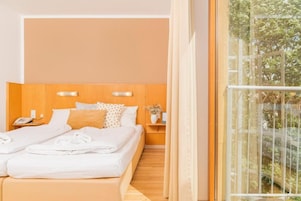 Quarto de hotel com cabeceira de madeira, cama de casal com lençóis brancos, mesa de cabeceira com telefone e vista para o verde pela janela.