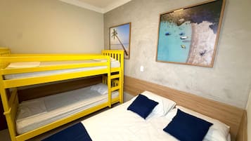 Um quarto com beliches e uma cama simples, um quadro com cena de praia e uma parede sem decoração.