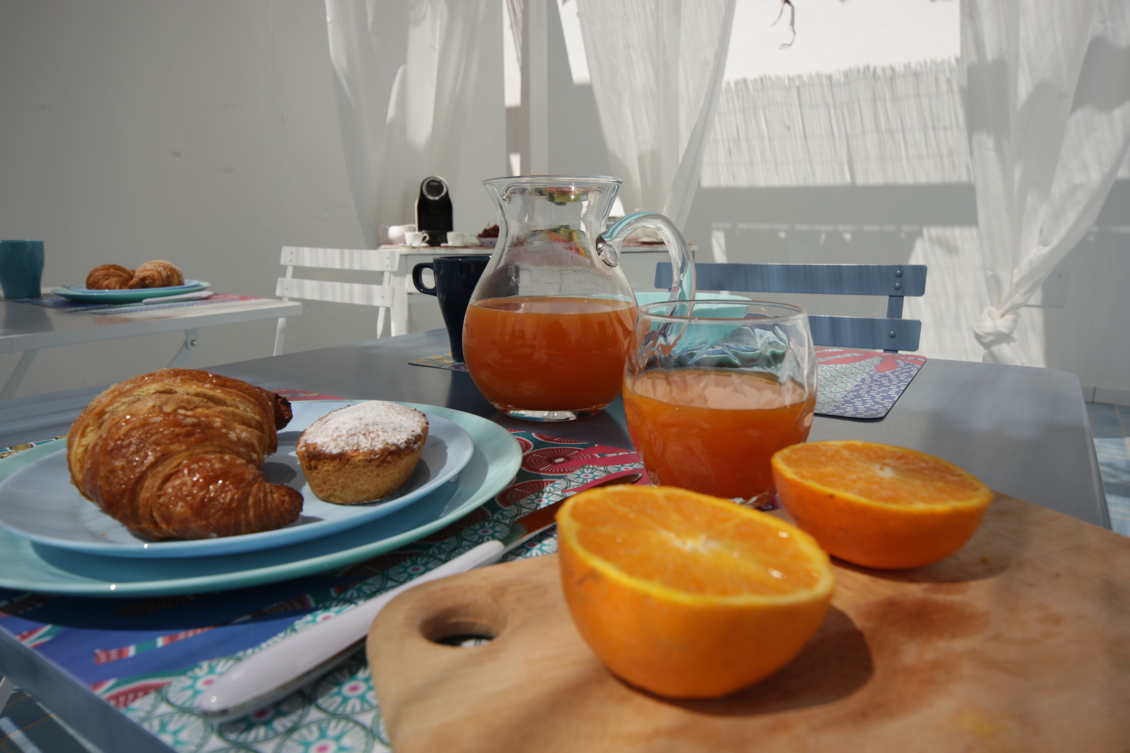 Mesa de café da manhã com croissants, muffins, suco de laranja e um jarro de chá sobre toalha de mesa estampada.