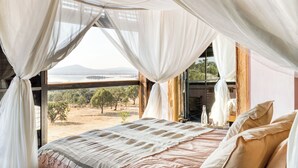 Quarto com uma grande janela, cama com roupa de cama branca e vista para uma paisagem natural.