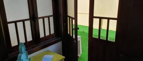 Quarto com porta de madeira, uma pequena mesa com um objeto azul e vista para um gramado verde lá fora.