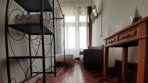 Quarto com piso de madeira em padrão herringbone, estante de metal preta, mesa de cabeceira de madeira com gavetas, sofá preto e janela com cortinas leves.