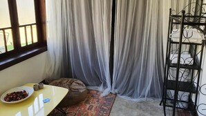 Quarto com cortinas leves, uma estante de metal, uma mesinha com uma tigela de frutas e um tapete estampado.