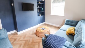 Sala de estar com um sofá azul, um banquinho redondo de madeira e uma televisão fixada em uma parede azul escura.