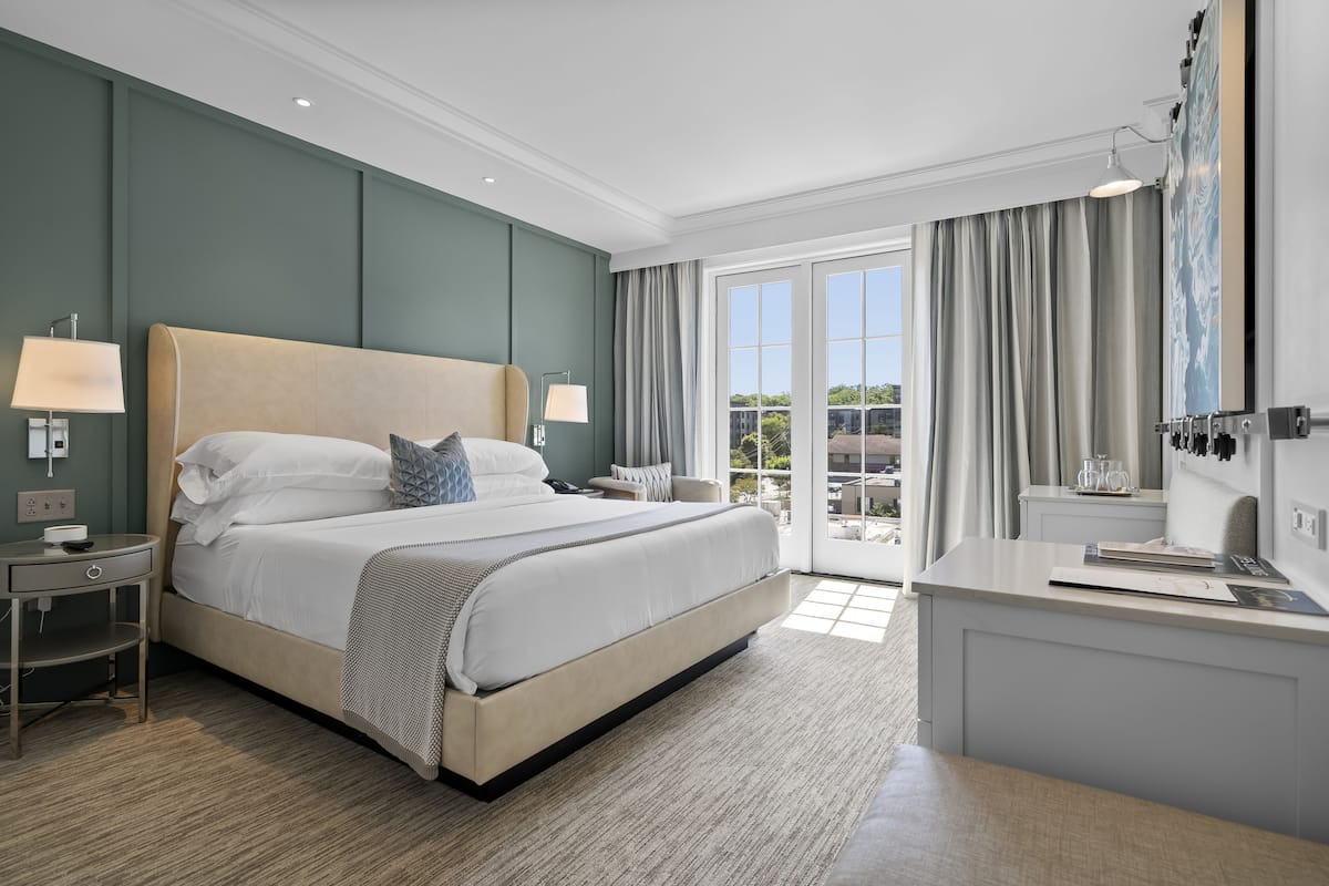 Quarto de hotel moderno com uma cama grande, mesas de cabeceira, escrivaninha e vista para o exterior.