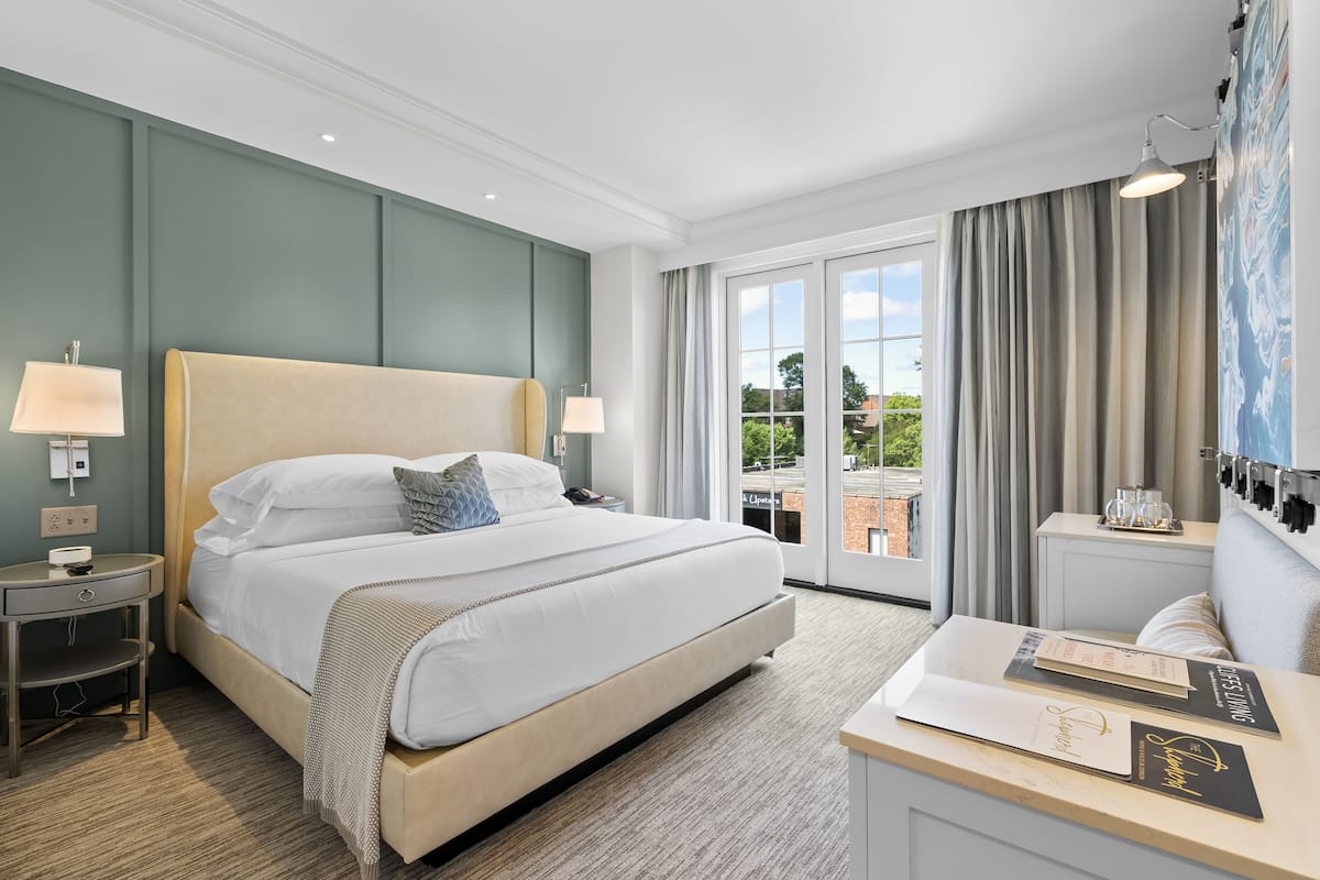 Quarto de hotel com uma cama grande, uma escrivaninha com luminária e vista para o exterior por uma porta de correr.