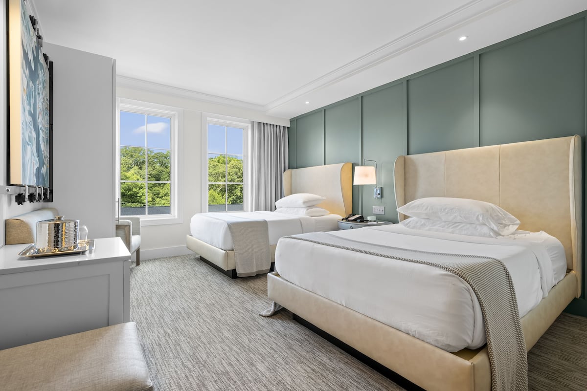 Quarto de hotel com duas camas, uma escrivaninha e uma janela grande com vista para o verde.