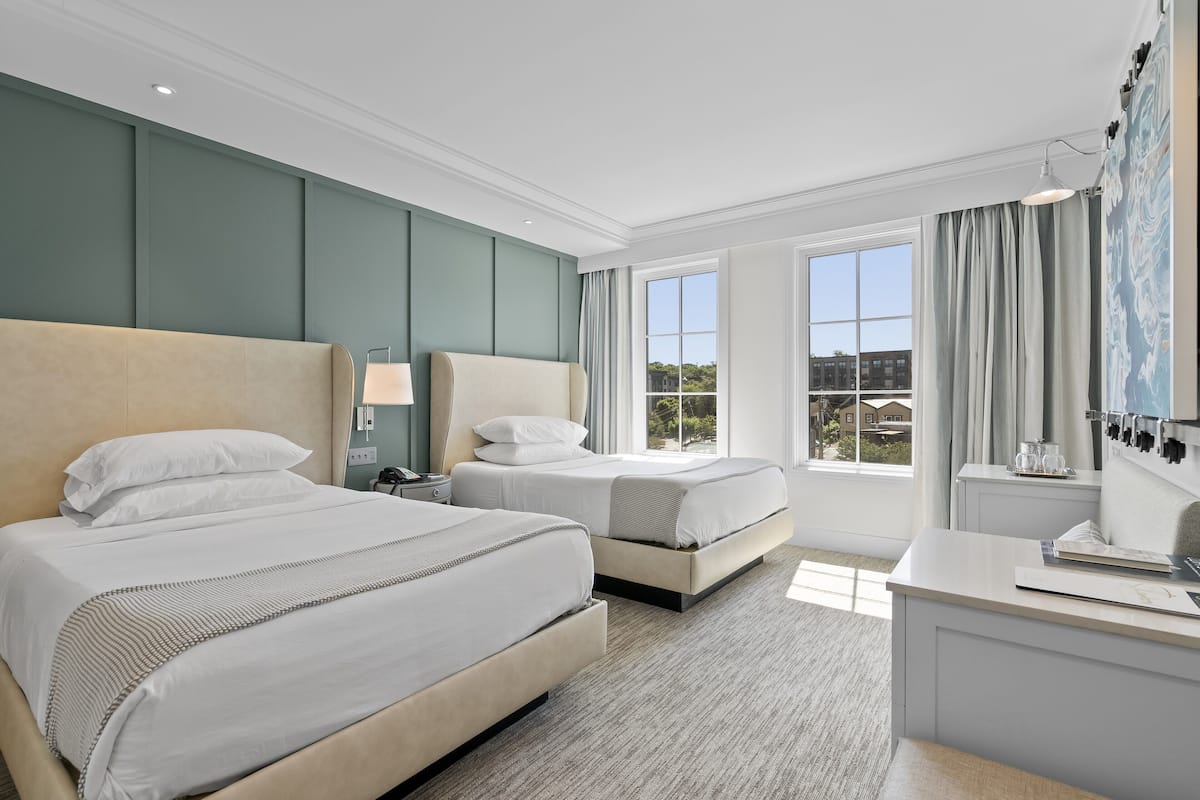 Quarto de hotel com duas camas, uma escrivaninha, uma luminária e vista para a cidade.