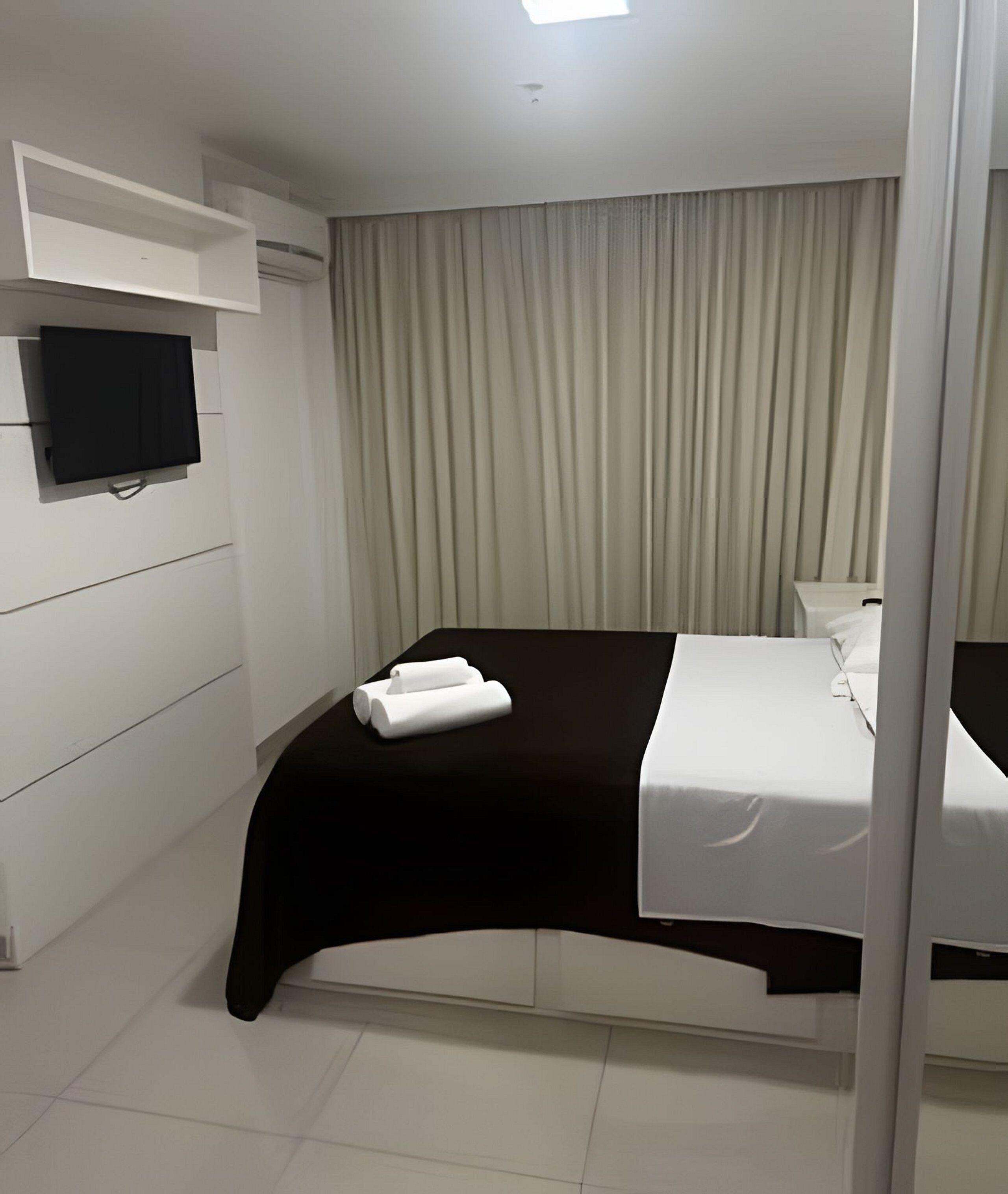 Quarto de hotel com cama, televisão de tela plana na parede e janela com cortinas.