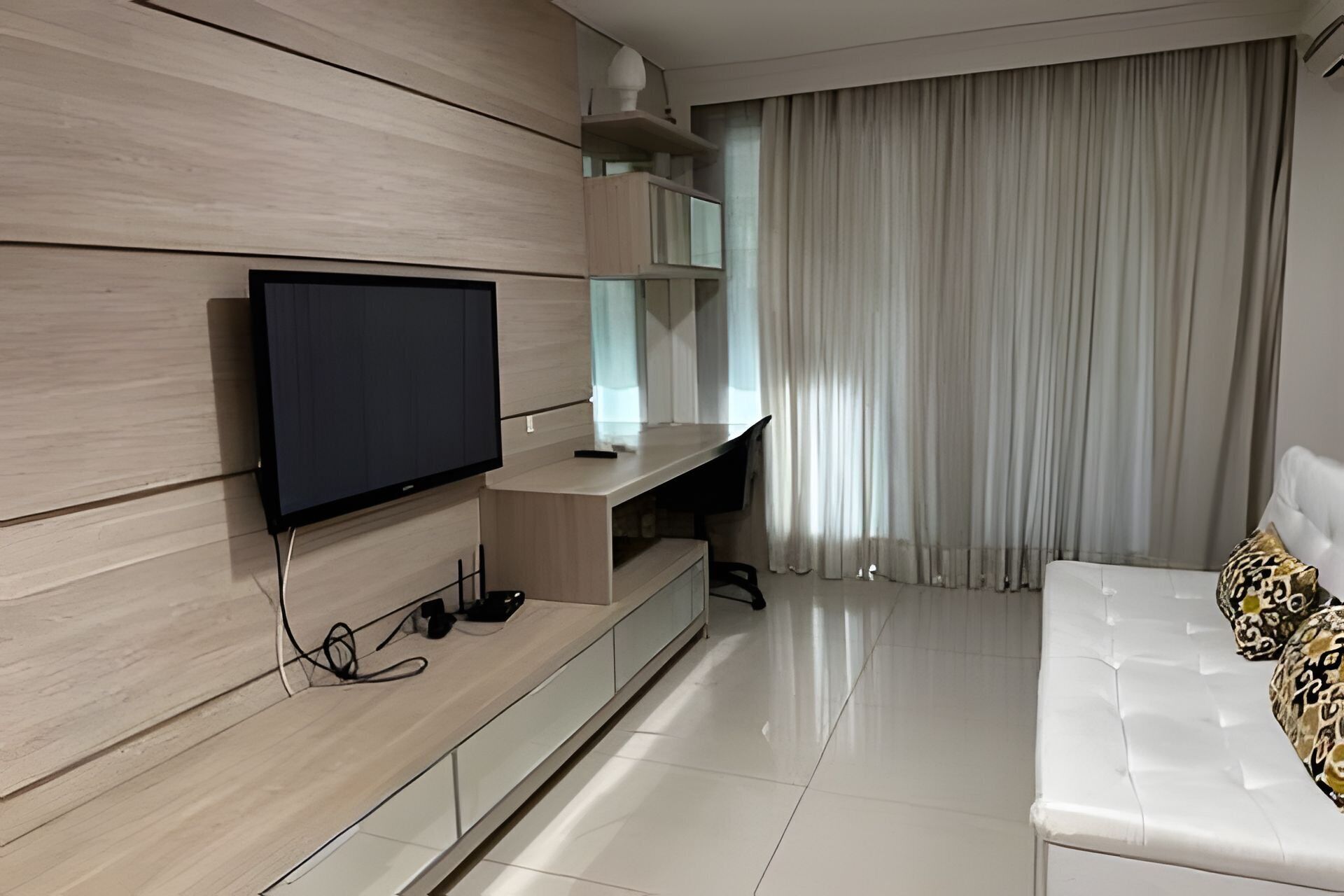 Uma sala de estar moderna com uma televisão de tela plana fixada em uma parede de madeira, um sofá branco com almofadas estampadas e uma grande janela com cortinas leves.