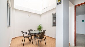Um pequeno espaço de pátio fechado com uma mesa redonda e quatro cadeiras, um vaso de planta e uma porta que dá para outro cômodo.
