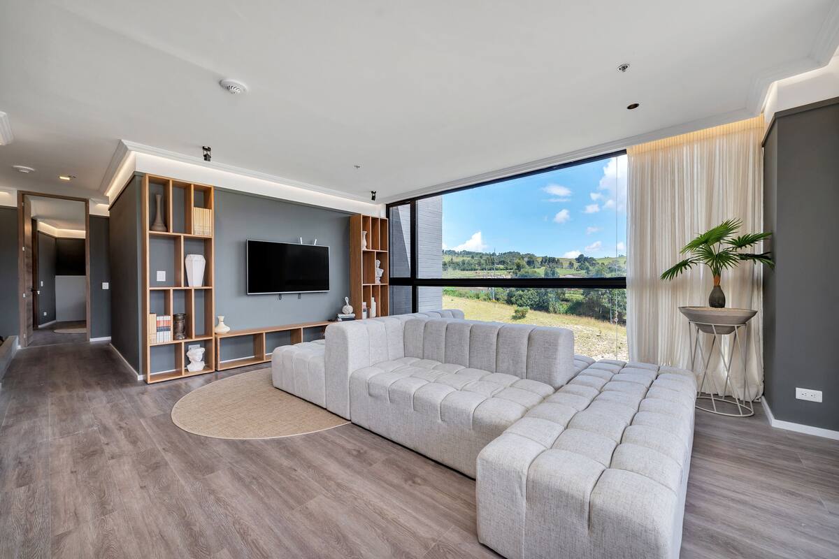 Uma sala de estar moderna com um amplo sofá modular, uma televisão de tela plana e vista para uma paisagem verde através de grandes janelas.