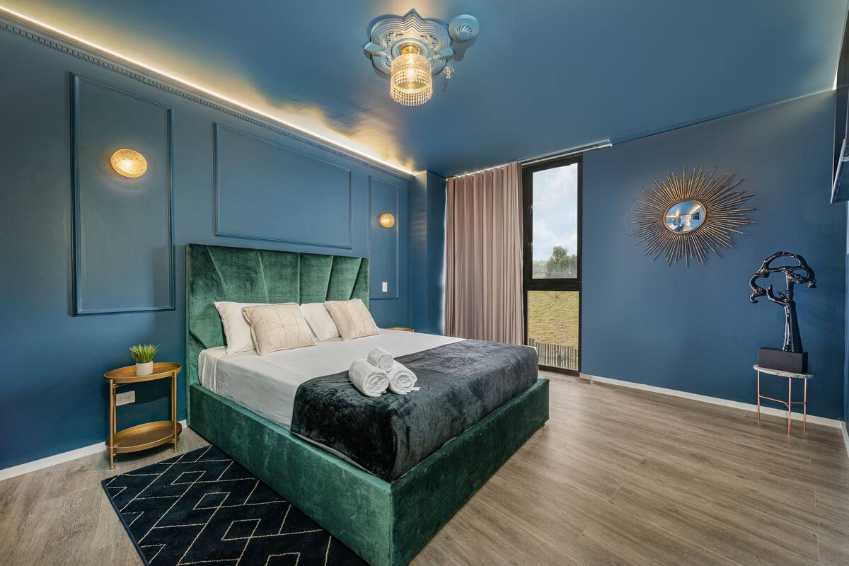 Quarto com cama grande, mesa de cabeceira, lustre e vista para o exterior.