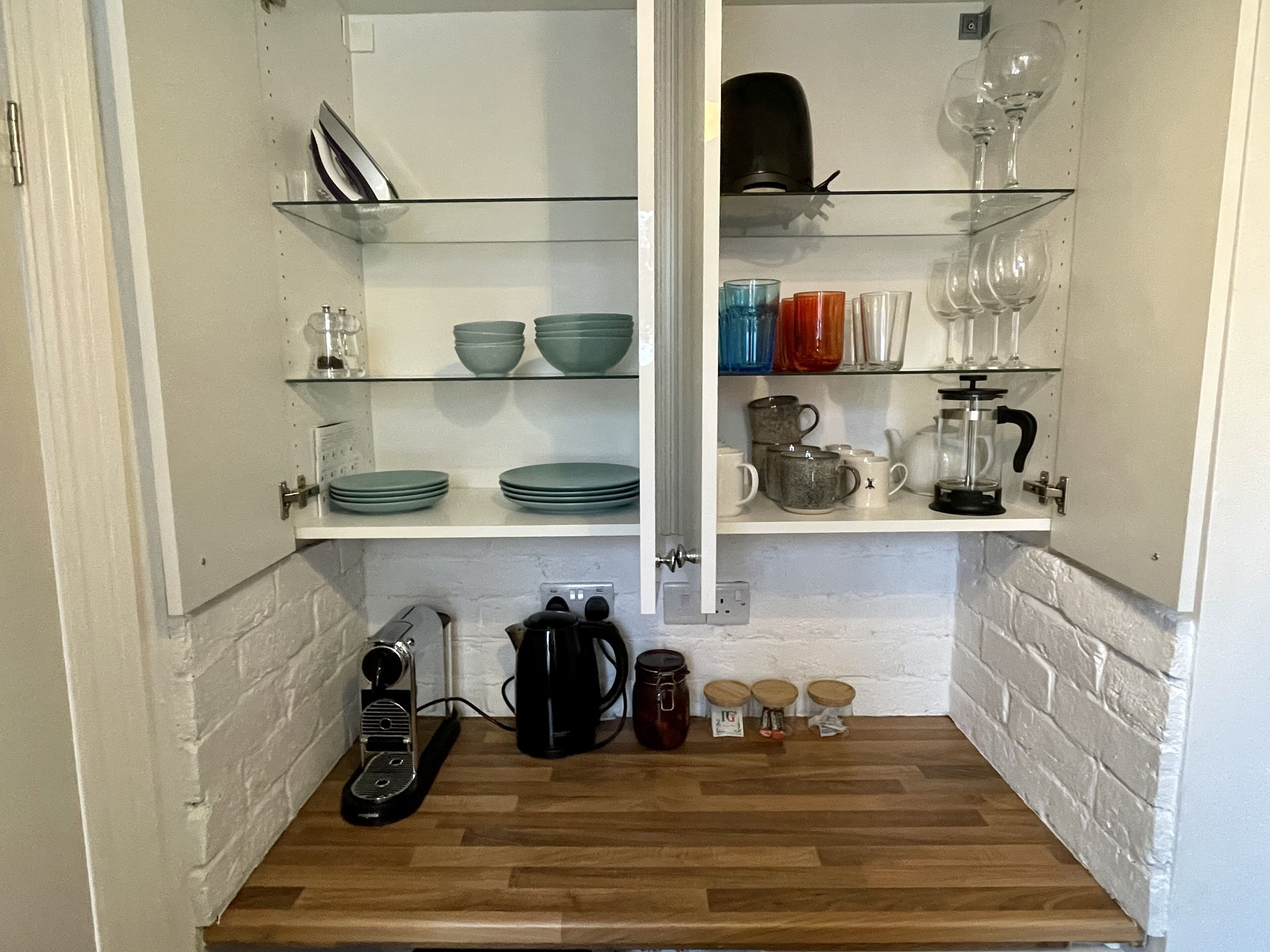 Uma cozinha com bancadas de madeira, prateleiras de vidro e diversos utensílios, incluindo pratos, tigelas e copos.