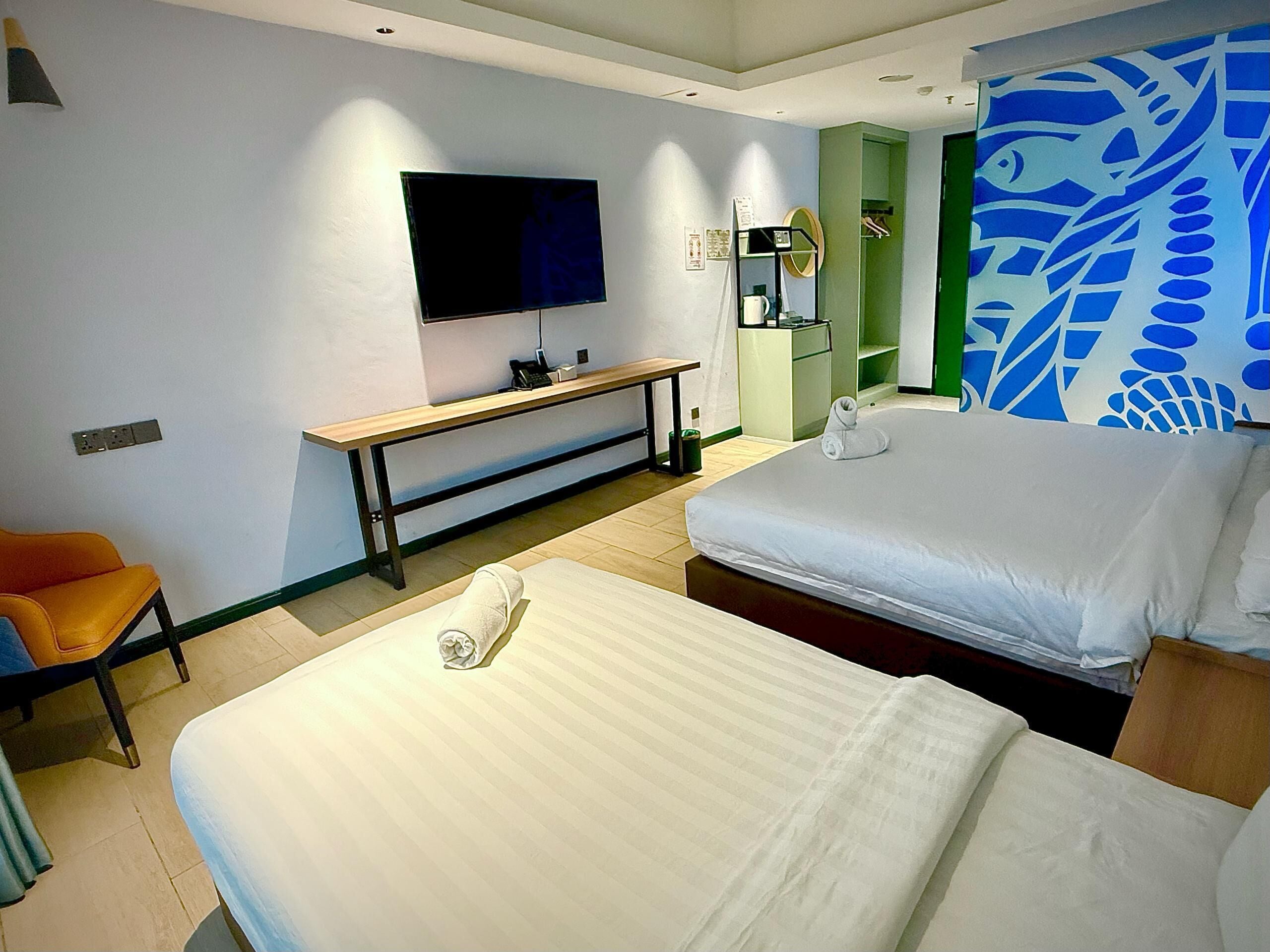 Quarto de hotel com duas camas, uma escrivaninha, uma televisão e uma parede verde com padrão azul.
