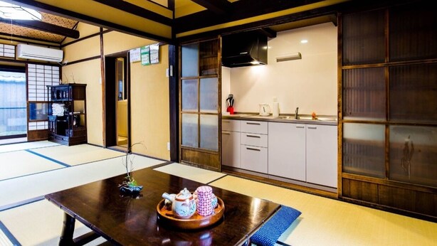 Um quarto japonês tradicional com tatames, uma mesa baixa com um conjunto de chá, uma cozinha com fogão e armários, e portas deslizantes.