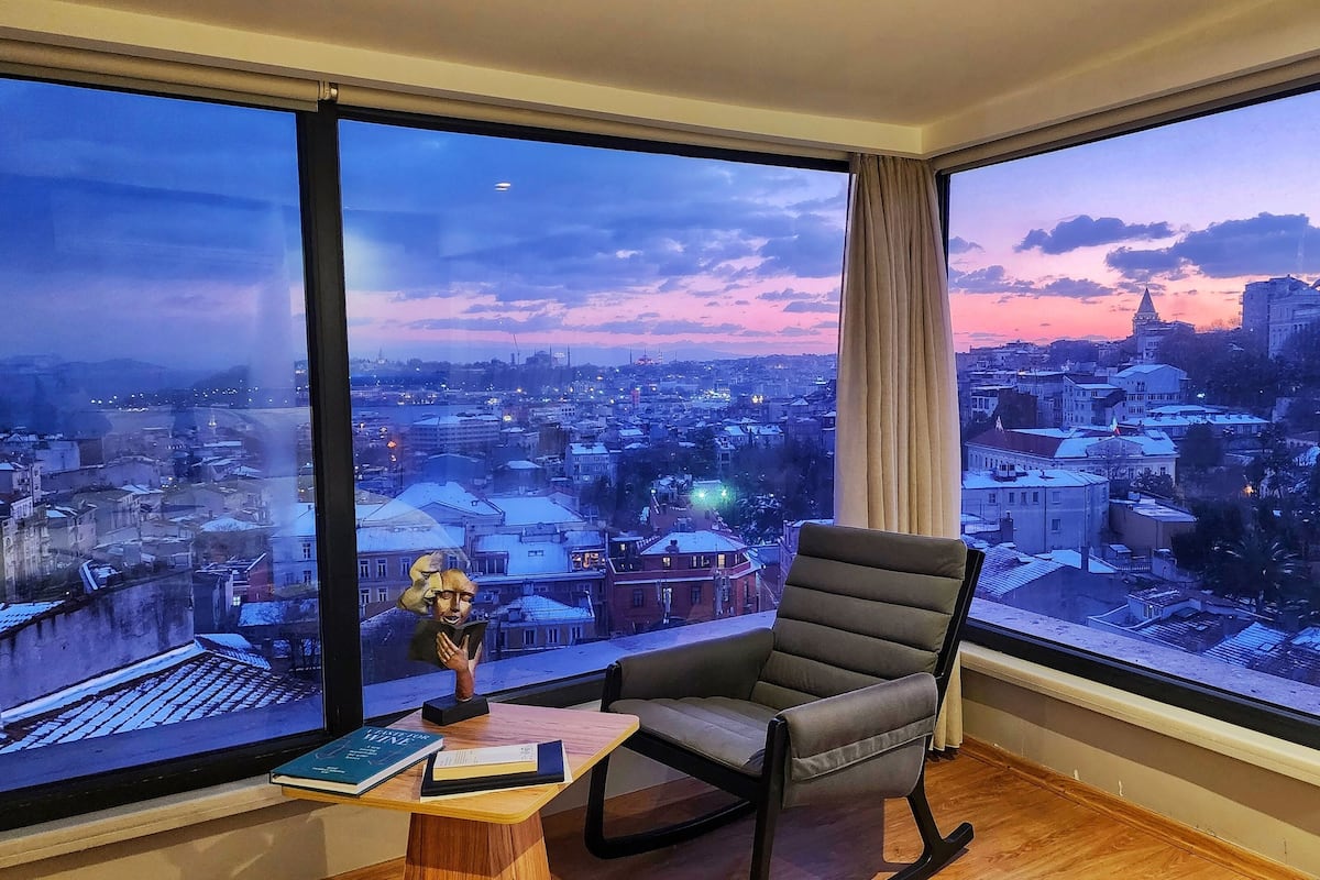 Quarto moderno com uma grande janela que oferece vista da cidade ao entardecer.