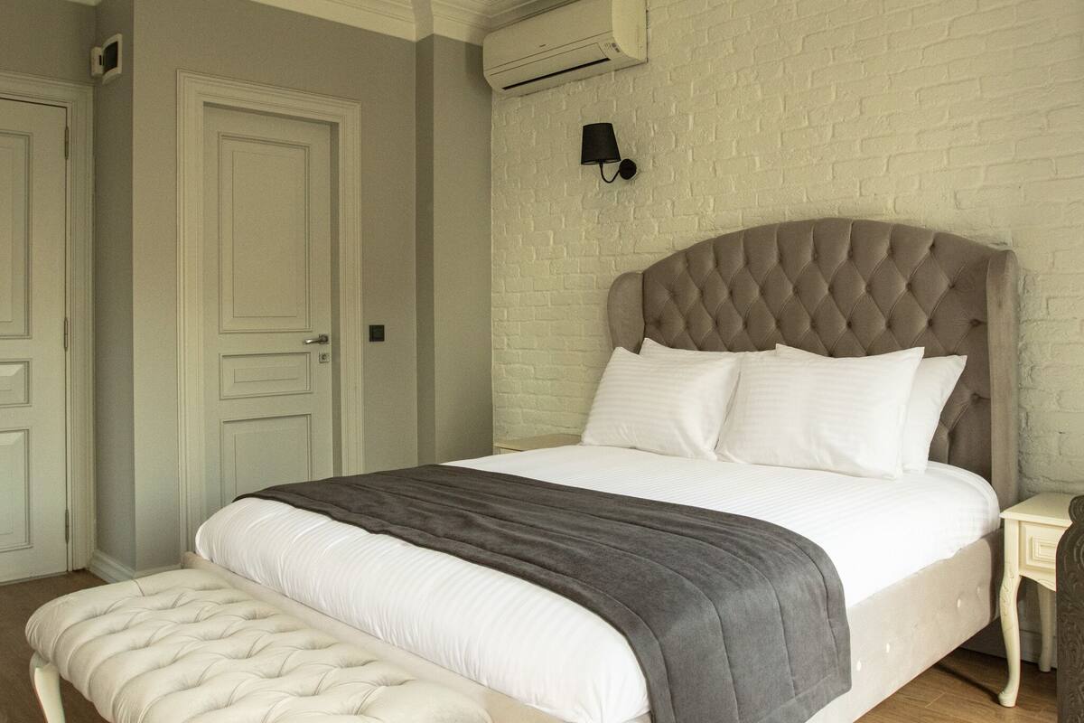 Quarto com uma cama grande, um banquinho, uma mesinha de cabeceira e uma parede de tijolos.
