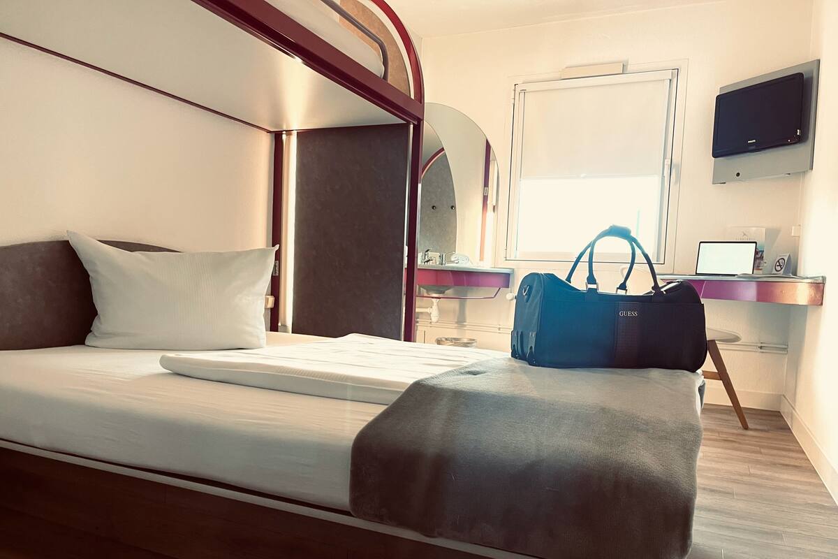 Quarto de hotel com uma cama, uma escrivaninha com computador, uma televisão e uma janela.