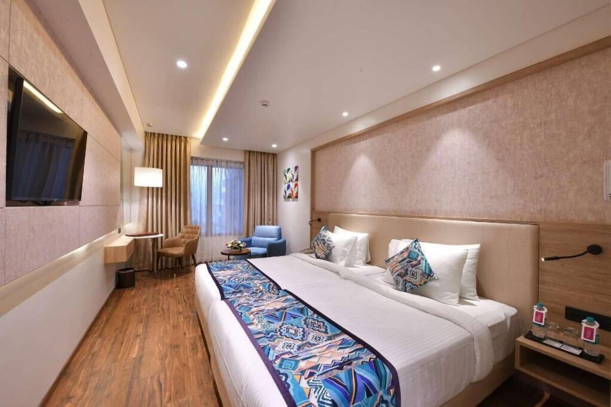 Quarto de hotel com uma cama grande, uma mesa com cadeira, TV e janela com cortinas.