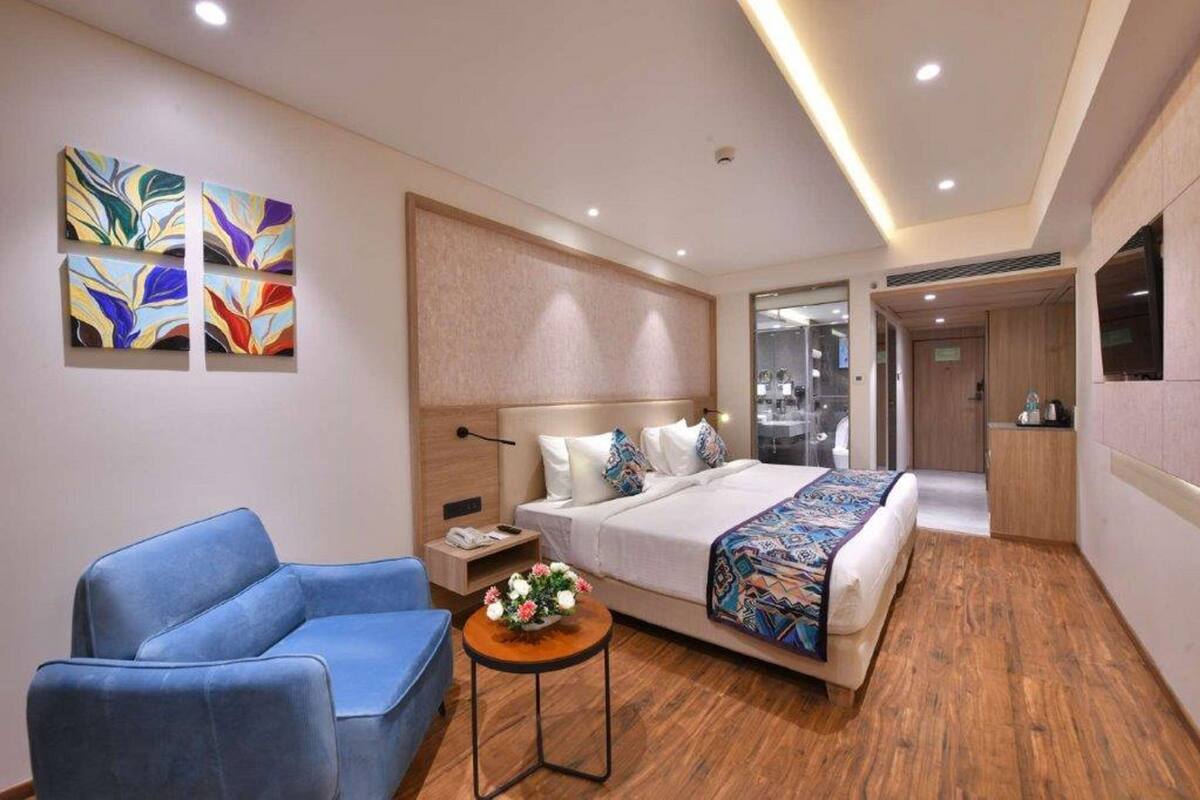 Quarto de hotel moderno com uma cama grande, uma poltrona azul, uma pequena mesa com flores e arte abstrata na parede.