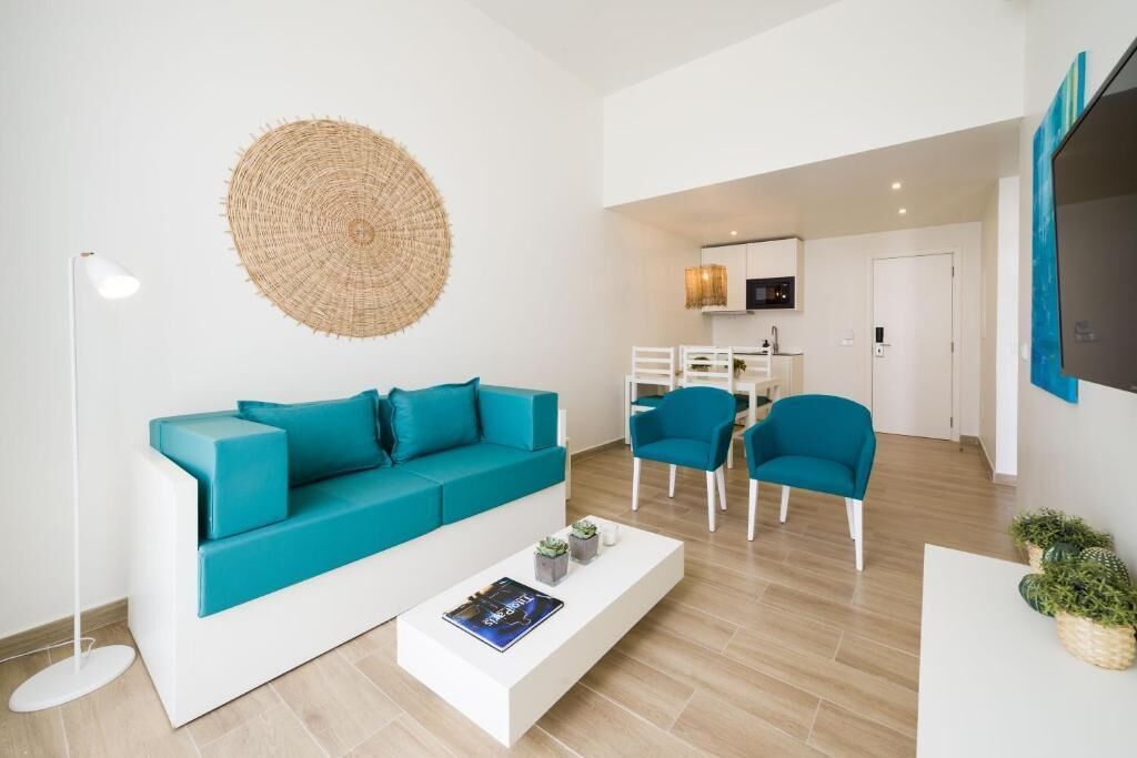 Uma sala de estar moderna com um sofá verde-água, mesa de centro branca e cadeiras a combinar. Uma peça de arte de parede trançada e uma televisão de tela plana são visíveis.