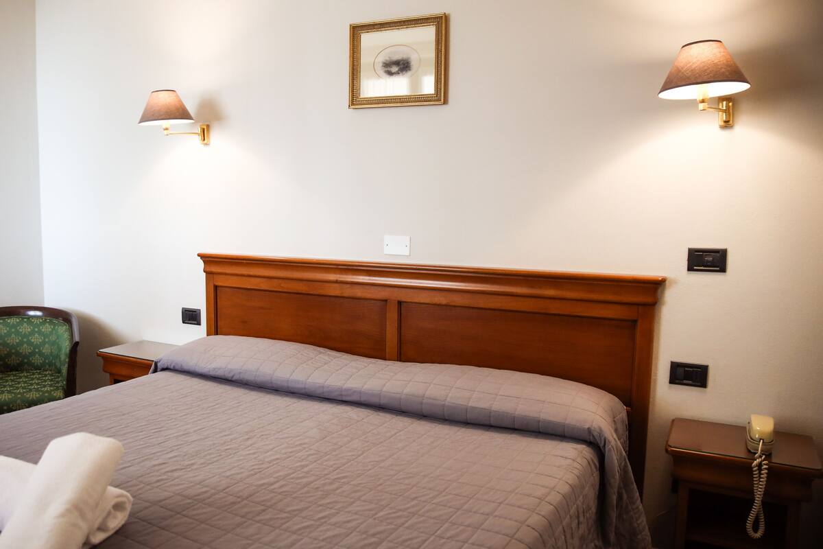 Quarto de hotel com cabeceira de madeira, cama com colcha acolchoada, poltrona estofada verde, mesa de cabeceira com abajur e telefone.