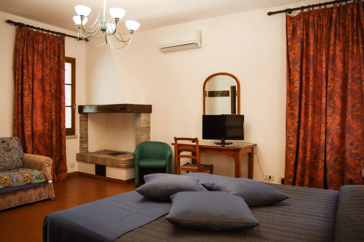 Quarto com sofá, cama com almofadas, escrivaninha com computador, lareira, espelho e cortinas.
