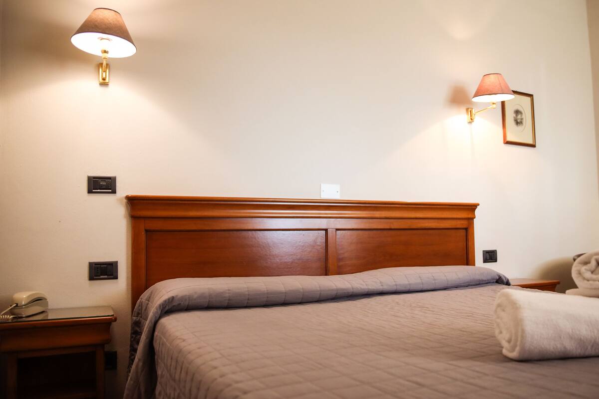 Quarto de hotel com cabeceira de madeira, cama com colcha acolchoada, duas luminárias fixadas na parede, mesa de cabeceira com telefone e toalha enrolada na cama.