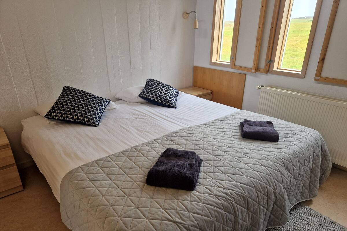 Cama bem arrumada com um edredom cinza, dois travesseiros estampados e duas toalhas dobradas.