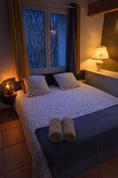 Quarto com cama, almofadas, abajur, janela com cortinas e vista para árvores.