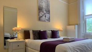 Quarto de hotel com uma cama grande, duas mesas de cabeceira com luminárias, um espelho, um quadro na parede e uma janela.
