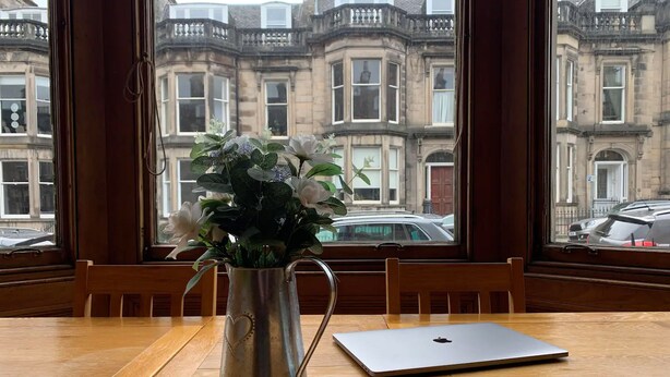 Uma mesa de madeira com um laptop e um jarro de metal com flores.