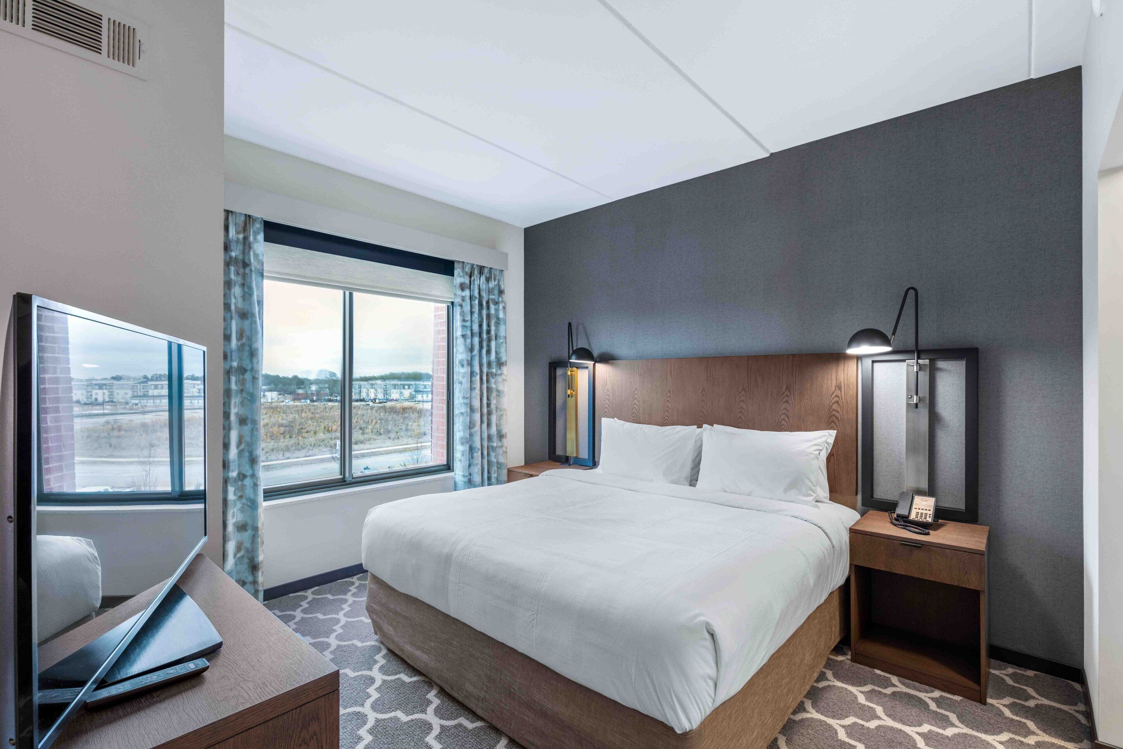 Quarto de hotel com uma cama grande, uma mesa com um laptop, uma mesa de cabeceira com uma luminária e vista para a cidade pela janela.