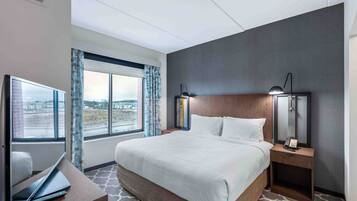 Quarto de hotel com uma cama grande, uma mesa com um laptop, uma mesa de cabeceira com uma luminária e vista para a cidade pela janela.