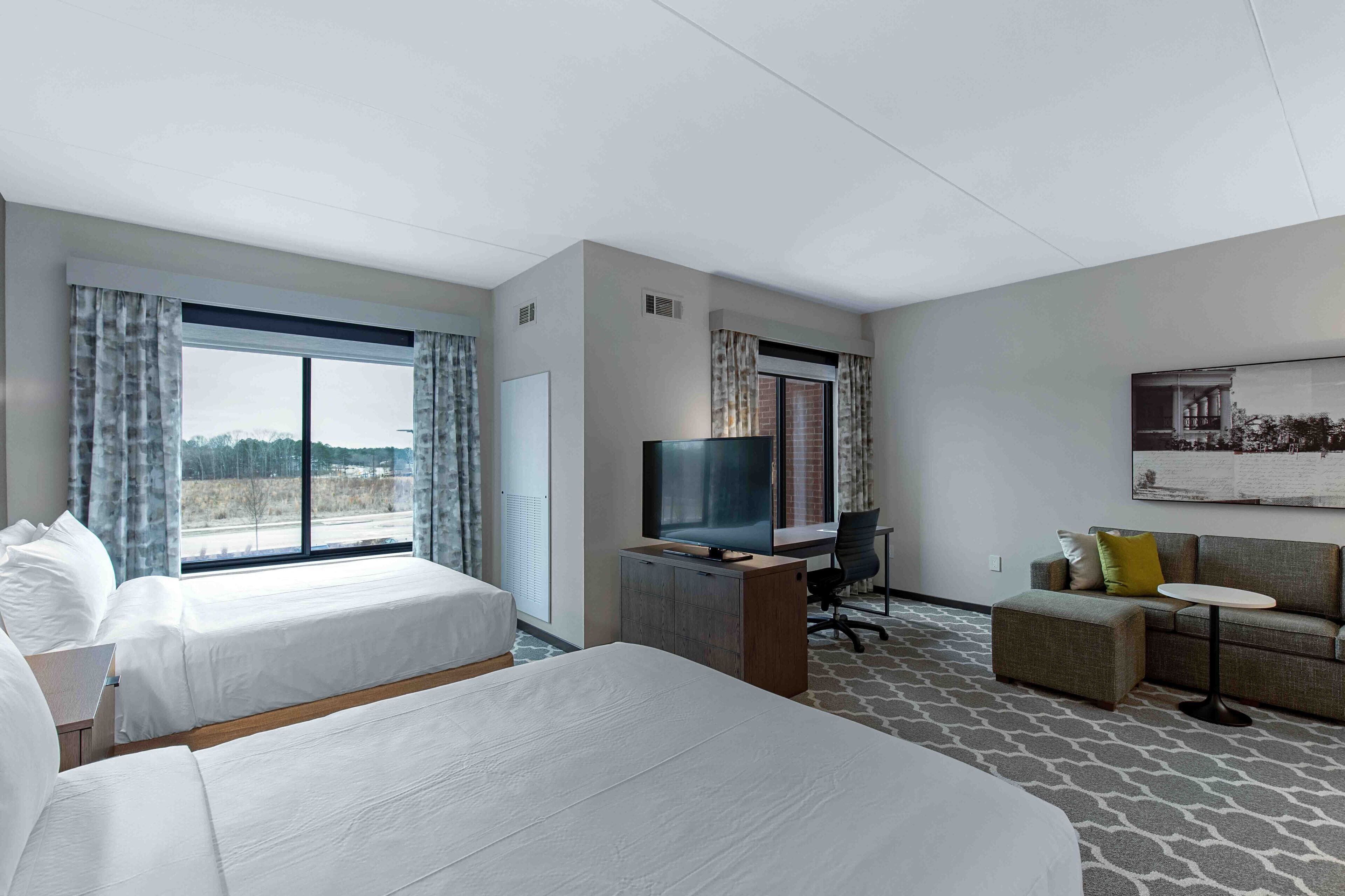 Quarto de hotel com duas camas, uma escrivaninha com televisão, um sofá e uma mesinha.