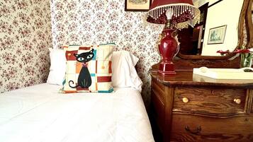 Quarto com papel de parede floral, cama com travesseiro xadrez, cômoda de madeira com espelho e abajur vermelho.