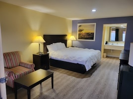 Quarto de hotel com cama, abajures, sofá, mesa, pintura, espelho e uma pequena cozinha equipada.