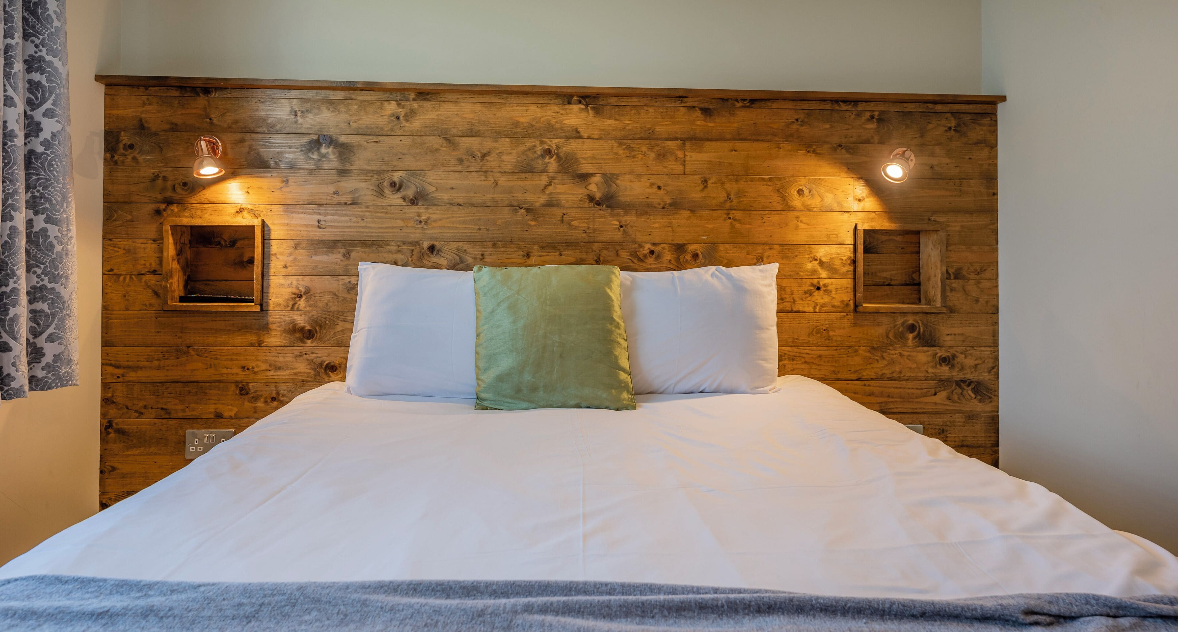 Cama com roupa de cama branca e um travesseiro verde, apoiada em cabeceira de madeira com duas luzes fixadas na parede.
