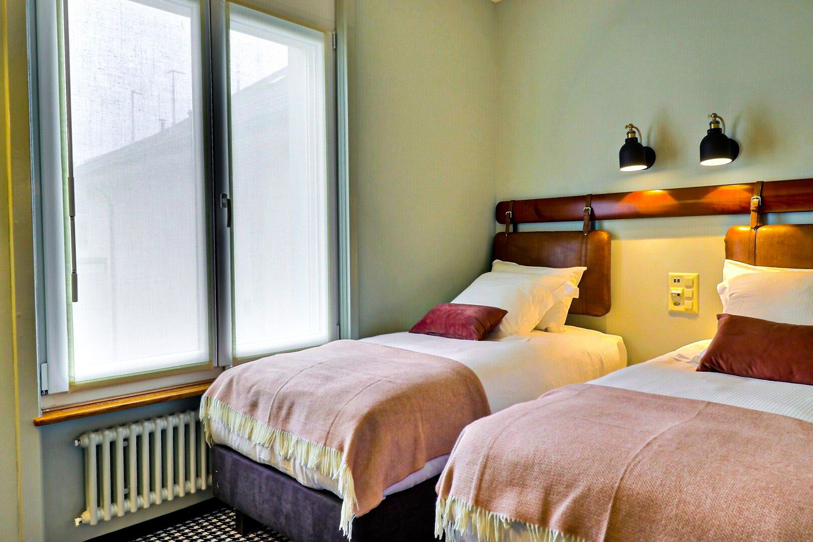 Quarto de hotel com duas camas, uma janela, um radiador e duas luminárias fixadas na parede.