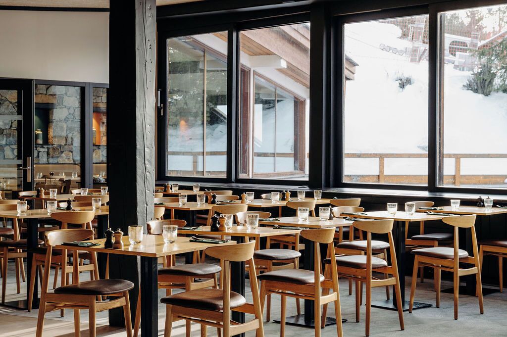Um restaurante com mesas e cadeiras de madeira, grandes janelas com vista para uma paisagem coberta de neve.