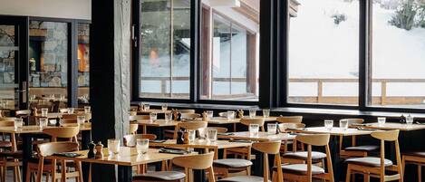 Um restaurante com mesas e cadeiras de madeira, grandes janelas com vista para uma paisagem coberta de neve.
