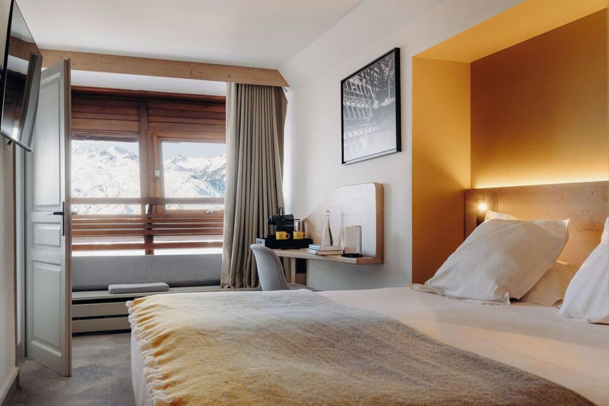 Quarto de hotel com cama, escrivaninha, cadeira, televisão, janela com cortinas e um quadro na parede.